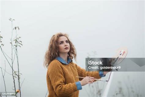 야외에서 그림을 그리는 젊은 여성 예술가 그녀는 캔버스와 그림 앞에 있습니다 그녀는 오일 페인트 아티스트 브러쉬 캔버스 및 팔레트를 보유하고 있습니다 곱슬 머리를 가진 농축