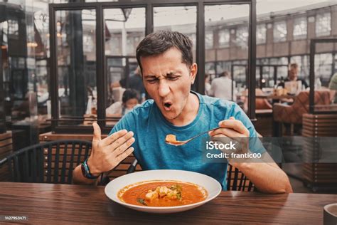한 남자가 식당에서 매콤하고 뜨거운 붉은 수프를 먹고 감정적으로 우스꽝스럽게 반응합니다 국가 요리의 조미료와 고추가 과다한