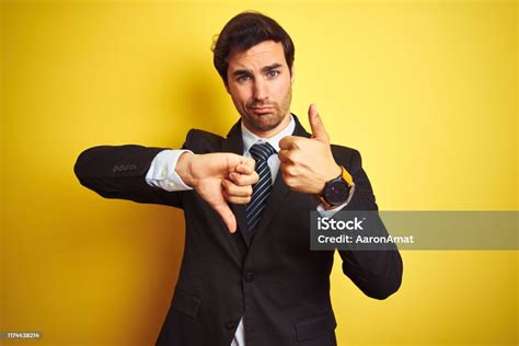 양복과 넥타이를 입고 고립된 노란색 배경 위에 서있는 젊은 잘생긴 사업가 엄지 손가락 위아래로 불일치및 계약 표현 미친 충돌