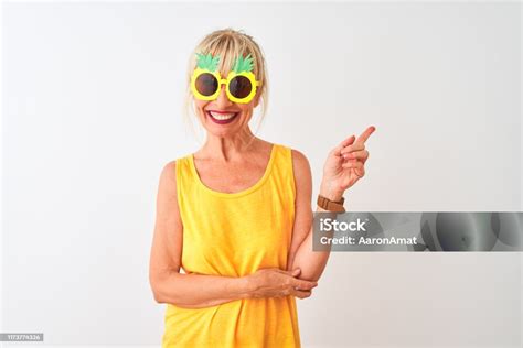 고립된 흰색 배경 위에 파인애플 선글라스를 쓰고 있는 중년 여성이 얼굴에 큰 미소를 지으며 카메라를 바라보며 손과 손가락으로 가리키는 모습 가리키기에 대한 스톡 사진 및