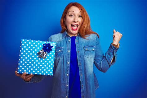 젊은 아름다운 빨간 머리 여자 입고 생일 선물 위에 고립 된 파란색 배경 비명 자랑과 승리와 성공을 축하 매우 흥분 응원 감정 감정에 대한 스톡 사진 및 기타 이미지