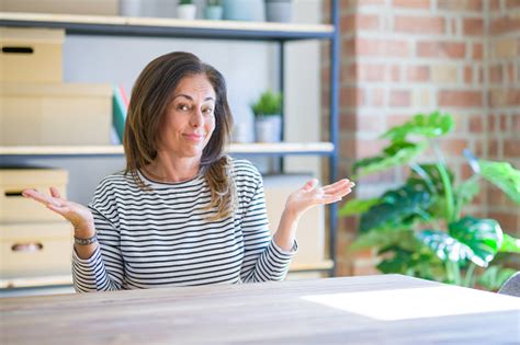 집에서 테이블에 앉아 있는 중년 노인 여성은 팔과 손을 들고 단서가 없고 혼란스러운 표정을 짓고 있습니다 의심 개념 가정 생활에