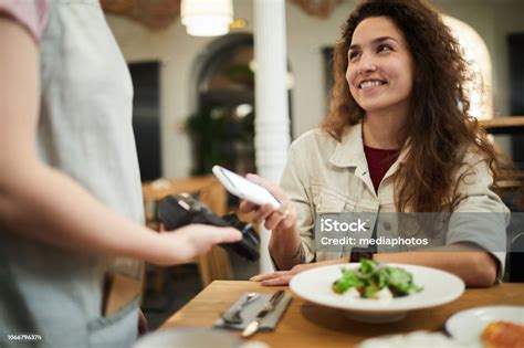 테이블에 앉아 하 고 스마트폰 터미널에 카페에 지불을 위한 Nfc 기술을 사용 하는 동안 곱슬 머리를 가진 쾌활 한 젊은 혼합된 인종 여자의 근접 촬영 지불에 대한 스톡