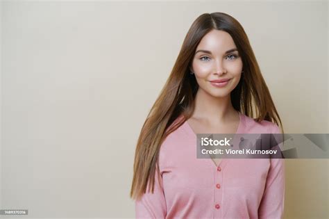 매력적인 유럽 여성의 수평 초상화는 카메라에서 부드럽게 미소 긴 직선 머리를 가지고 캐주얼 장미 스웨터를 입고 야외 산책을하려고 베이지 색 스튜디오 배경에 대해 서있다 25