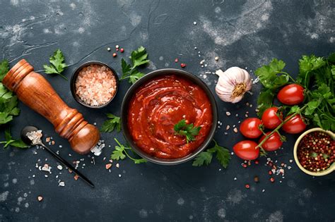 토마토 케첩 매운 소스와 재료 파슬리 마늘 후추는 어두운 슬레이트 돌 또는 콘크리트 배경에 향신료와 신선한 토마토와 검은 그릇에 복사 공간이 있는 상단 뷰입니다 가벼운에