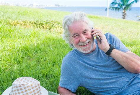 전화로 말하는 흰 머리와 수염을 가진 쾌활한 노인 잔디에 누워 컴퓨터에서 작업 손에 전화 배경의 푸른 바다 공원에 대한 스톡 사진 및 기타 이미지 Istock