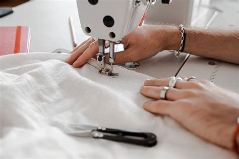 바느질 기계 근처의 남성 손은 봉제 봉합 과정에서 흰색 천을 잡고 있습니다 바느질 디자인 작업 재단 사 재단사 디자이너 의류 제조 소 창조적 인 작업 과정에서 가위에 대한