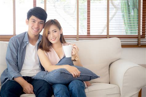 아름 다운 젊은 아시아 커플 미소와 이야기 이야기 결혼 집에서 소파에 앉아 함께 여자와 함께 커피 한 잔을 들고 남자와 여자