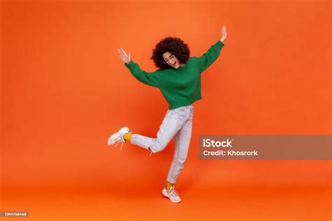 한쪽 다리에 서 녹색 캐주얼 스타일 스웨터를 입고 아프로 헤어 스타일을 가진 행복한 여성의 전체 길이 초상화 팔을 들어 올리고 춤을 추며 축하합니다 춤에 대한 스톡 사진 및