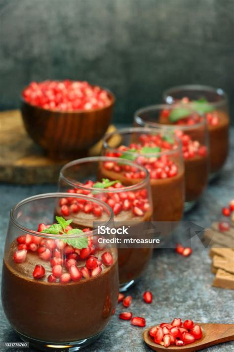 석류 씨앗과 민트 잎 장식을 얹은 홈메이드 초콜릿 무스 디저트가 들어 있는 대각선 안경의 이미지 밀크 초콜릿 덩어리 나무 숟가락 석류 씨앗의 나무 그릇 어두운 회색 배경