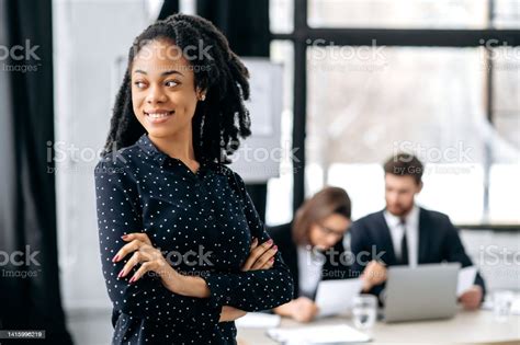 브리핑 회의에서 사랑스러운 우아한 젊은 비즈니스 여성의 초상화 성공적인 아프리카 계 미국인 여성 팔을 교차 한 창조적 인 현대 사무실에 서서 옆을 바라보고 미소 짓고