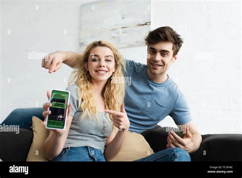 Selective Focus Of Happy Man And Blonde Woman Pointing With Fingers At Smartphone With Booking