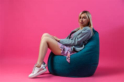 파란색 재킷에 아름다운 금발 소녀는 삼각형의 모양으로 떨어져 그녀의 다리와 핑크 고립 된 배경에 그들 사이에 접힌 손으로 녹색 가방 의자에 앉아 빈백에 대한 스톡 사진 및