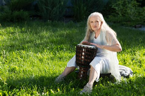 긴 회색 머리를 가진 행복한 노인 여성은 화창한 날 잔디밭에 앉아 야외에서 레크리에이션 활동이나 치료로 민족 드럼을 연주합니다 모두를 위한 지식 1960 1969 년에 대한