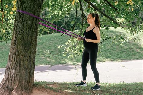 검은 색 점프 수트를 입은 운동하는 검은 머리의 임산부는 여름날 공원에서 체육관 탄성 밴드와 함께 운동을하고 있습니다 건강한 라이프 스타일의 개념 산전 관리 모성 및 임신