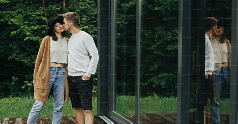 세련된 힙스터 커플은 나무 테라스에서 포옹하고 키스하며 산의 오두막에서 휴식을 취합니다 큰 창문과 나무를 배경으로 현대적인