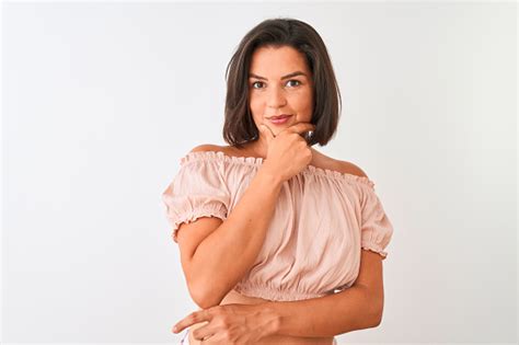 고립된 흰색 배경 위에 서있는 캐주얼 한 티셔츠를 입은 젊은 아름다운 여성은 턱에 손을 들고 교차 된 팔과 손으로 미소 짓는 카메라를 보고 자신감을 보입니다 긍정적 인 생각