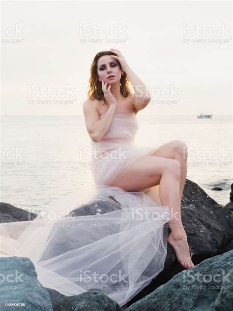 분홍색 얇은 명주 그물의 아름다운 모델은 석양의 광선에 바다 해안의 돌에 앉아 있습니다 매력적인 슬림 여성의 전체 길이 초상화 35 39세에 대한 스톡 사진 및 기타 이미지