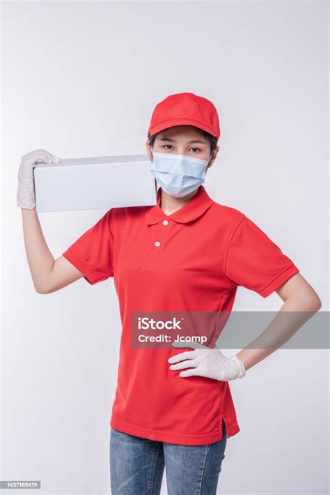 빨간 모자 빈 티셔츠 유니폼 얼굴 마스크 장갑을 낀 의식이 있는 젊은 배달원의 이미지는 밝은 회색 배경 스튜디오에 격리된 빈 흰색 판지 상자 팬데믹 코로나바이러스 바이러스