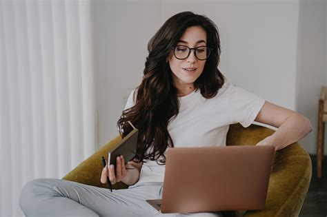 물결 모양의 머리카락을 가진 진지한 갈색 머리 여자가 흰색 티셔츠와 가벼운 바지와 안경을 입은 노트북으로 의자에 앉아 있습니다 집에서 원격 작업 화상 통화를 모의하십시오