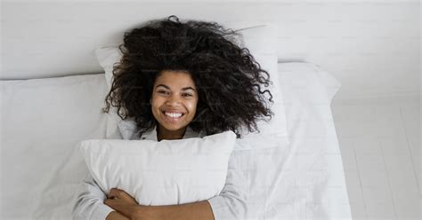 행복한 아프리카계 미국인 여성이 베개를 껴안고 활짝 웃으며 카메라를 바라보고 있는 모습 위 재미 있고 게으른 여자는 잠옷에 침대에 누워 집에서 이른 주말 아침을 보낸다