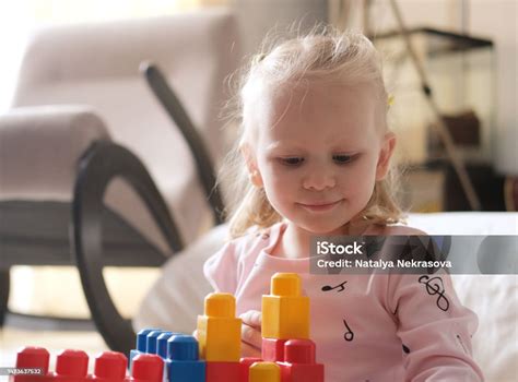 웃고 있는 어린 아이 화려한 큐브를 가지고 노는 금발의 미취학 아동 분홍색 잠옷을 입고 바닥에 앉아 집에 큰 창문이 있는 햇볕이 잘 드는 방 아이들의 초기 발달 2 3 살에