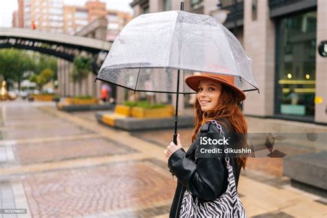 패션 모자를 쓰고 투명한 우산을 들고 야외에서 비오는 날씨를 즐기는 아름다운 도시 거리를 걷고 있는 웃고 있는 우아한 젊은 여성의 뒷모습 비에 대한 스톡 사진 및 기타