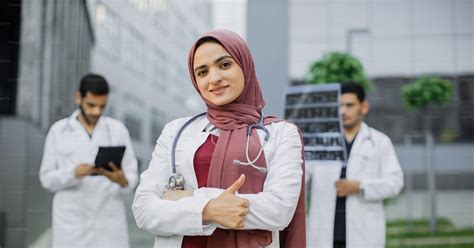 유니폼과 히잡을 쓴 아름다운 젊은 여성 아랍 의사가 카메라를 바라보며 엄지손가락을 치켜들고 웃고 있는 동안 그녀의 두 남자 자신감 넘치는 동료가 현대 병원 근처 야외에서