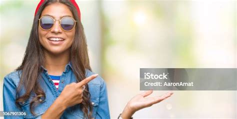 젊은 아름 다운 아랍 여자 놀라게 했 고 손으로 제시 하 고 손가락으로 가리키는 동안 카메라에 웃 고 고립 된 배경 위에 선글라스를 착용 가리키기에 대한 스톡 사진 및 기타