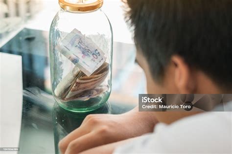 실망한 청년은 저축 돈이 있는 유리 항아리 콜롬비아 지폐 금융을 보고 있습니다 저축과 경제의 개념 낮은 경제 자원을 가진 가난한 소년 16 17 살에 대한 스톡 사진 및