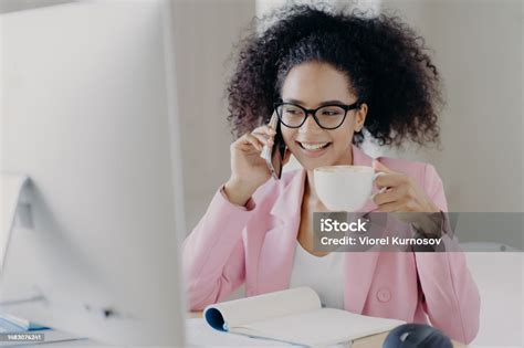 매력적인 미소를 짓고 있는 여성 사업가의 사진은 향기로운 음료를 즐기고 하얀 음료수 머그잔을 들고 전화 통화를 하고 정장을 입고 직장에서 포즈를 취하고 투명한 안경을 쓴다