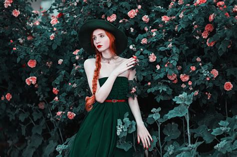 녹색 모자와 빨간 벨트를 가진 복장에 창백한 피부와 파란 눈 빨간 머리 소녀 부시 복숭아 장미 배경 긴 붉은 엮 다 가진 여자 빨간 입술 그의 목에 목걸이입니다 밝은 특이