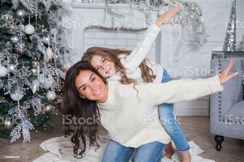새해 전야에 행복한 가족 젊은 아름다운 어머니와 흰색 아늑한 스웨터와 담요에 작은 귀여운 딸은 장식 된 크리스마스 트리 아래에서 재미와 포옹을합니다 축제 홈 클래식 인테리어