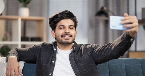 잘 생긴 수염을 기른 웃는 인도 남자 집에서 편안한 소파에 편안하게 앉아 스마트 폰을 사용하여 자기 사진을 찍거나 화상 통화를합니다 집에서 여가 가제트 개념 사진