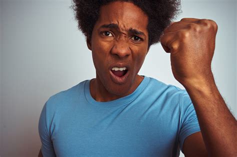 고립 된 흰색 배경 위에 서있는 파란색 티셔츠를 입은 아프리카 계 미국인 남성이 분노로 짜증나고 미친 듯이 외치며 손을 들고