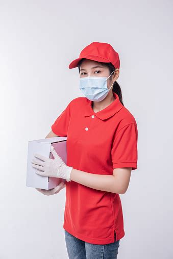 빨간 모자 빈 티셔츠 유니폼 얼굴 마스크 장갑을 낀 의식이 있는 젊은 배달원의 이미지는 밝은 회색 배경 스튜디오에 격리된 빈 흰색 판지 상자 팬데믹 코로나바이러스 바이러스