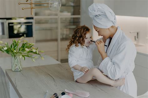 행복한 유럽 어머니는 딸 코에 화장품 브러시를 유지 흰색 부드러운 목욕 가운을 입고 얼굴 분말을 적용 외모와 피부에주의 가족의 아름다움 웰빙 어린 시절 모성 2 3 살에