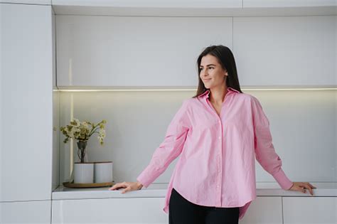 분홍색 셔츠와 검은 바지를 입은 자신감 넘치는 예쁜 주부가 부엌에 서서 귀여운 미소로 옆을 바라보며 건강하고 성공한 행복을 느낍니다 집에서 휴가를 꿈꾸는 느슨한 머리를 가진