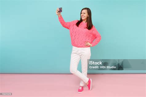 니트 장미 스웨터 흰색 바지 밝은 분홍색 파란색 파스텔 벽 배경에 고립 된 핸드폰에 촬영을 하 고 여자의 전체 길이 초상화 패션 라이프 스타일 개념입니다 복사 공간을 모의