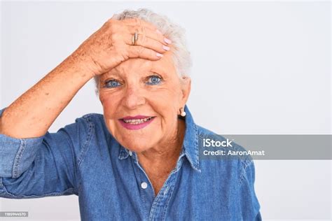 머리에 손으로 스트레스를 받은 고립된 흰색 배경 위에 서 있는 캐주얼 한 데님 셔츠를 입은 선임 회색 머리 여성은 수치심과 놀라움의 얼굴로 충격을 받았으며 화가 났습니다