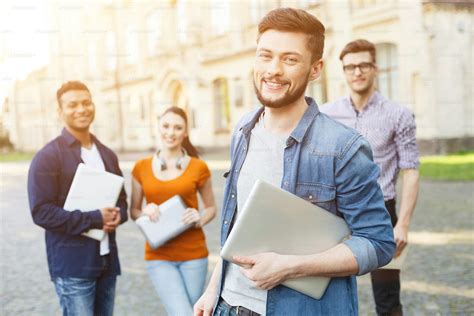 노트북을 들고 있는 잘생긴 청년의 초상화 그는 기대하고 웃고 있습니다 그의 친구들은 배경에서 대학에 대항하고 있습니다 사진 Unsplash의 기쁨 이미지