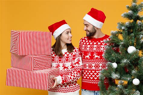 빨간 스웨터 크리스마스 모자에 미소 젊은 산타 커플 친구 남자 여자는 노란색 배경 스튜디오에서 고립 된 선물 전나무 나무와 선물 상자를 개최합니다 새해 축하 메리 홀리데이