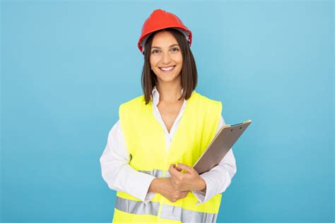 안전 조끼와 안전모를 착용하고 푸른 배경에 클립 보드를 들고 웃고있는 전문 여성 엔지니어 20 29세에 대한 스톡 사진 및 기타 이미지 20 29세 개성 개념 건설