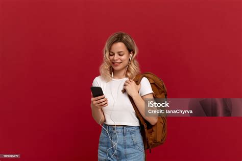 빨간 배경에 서 흰색 티셔츠를 입고 곱슬 금발 머리를 가진 여자의 초상화 오렌지 색 배낭과 흰색 헤드셋행복 모델은 전화기 화면에서 보인다 여행의 개념 T 셔츠에 대한 스톡