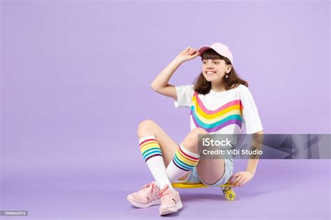 노란색 스케이트 보드에 앉아 스튜디오에서 보라색 파스텔 벽 배경에 고립 된 찾고 선명한 옷에 꽤 즐거운 십 대 소녀 사람들은 진심으로 감정 라이프 스타일 개념입니다 복사