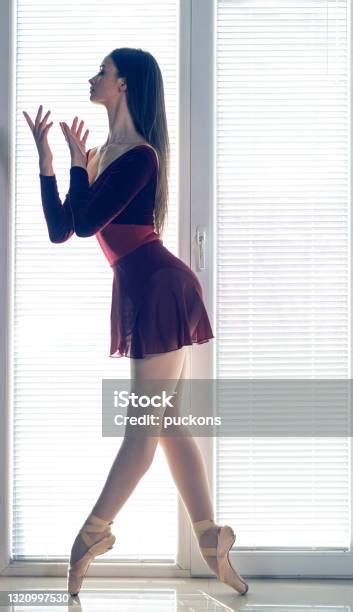 발레 스튜디오의 큰 창문 근처에서 진홍색 쇼트 드레스를 입고 포즈를 취하는 아름다운 젊은 발레리나 스탠드와 포즈를 취하고 있으며 자연채광은 블루 소프트 앰비언트 라이트로