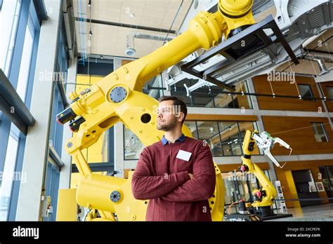Confident Engineer Standing With Crossed Arms In Front Of A Large Robotic Arm In A Modern