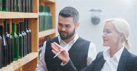 자신감 넘치는 금발 여성 소믈리에와 그녀의 동료가 레스토랑 지하실에서 병에 담긴 다양한 종류의 와인에 대해 토론하고 있다 사진 Unsplash의 동료 이미지