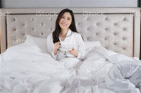 깨어난 후 아침에 침대에 앉아 있는 아시아 여성들 햇볕이 잘 드는 침실에서 침대에 어린 소녀 화창한 침실에서 따뜻한 커피를 즐기는 소녀를 미소 짓는 소녀 30 34세에 대한