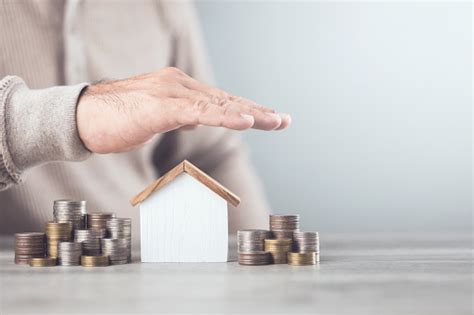 사업가가 동전 더미와 집 모형 위로 보호의 손짓을 하고 있습니다 이미지는 자산 보호의 개념을 나타내며 금융 투자 및 재산 보호를 강조합니다 계획에 대한 스톡 사진 및 기타